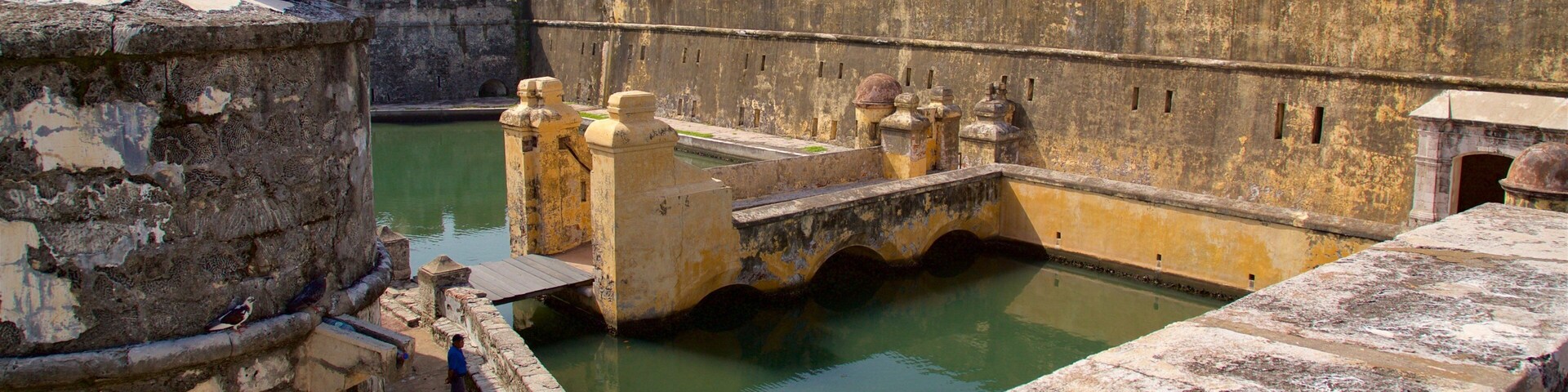 Uma fortaleza histórica com uma ponte sobre um fosso, rodeada por antigas muralhas de pedra e um edifício grande ao fundo.
