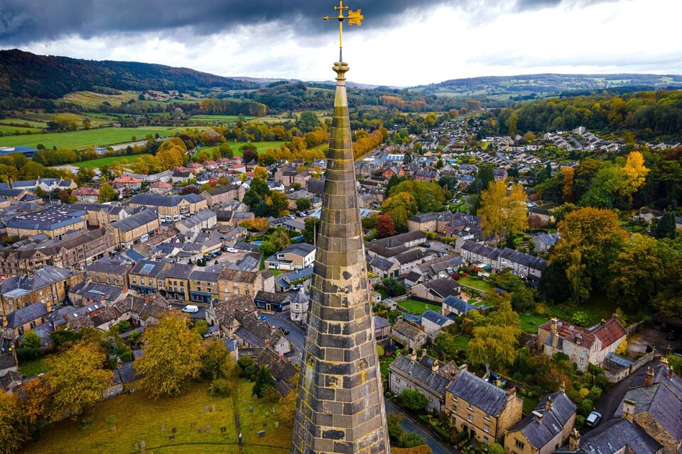 Uma torre histórica com um esguicho no topo, rodeada por uma aldeia com arquitetura tradicional e colinas ondulantes ao fundo.