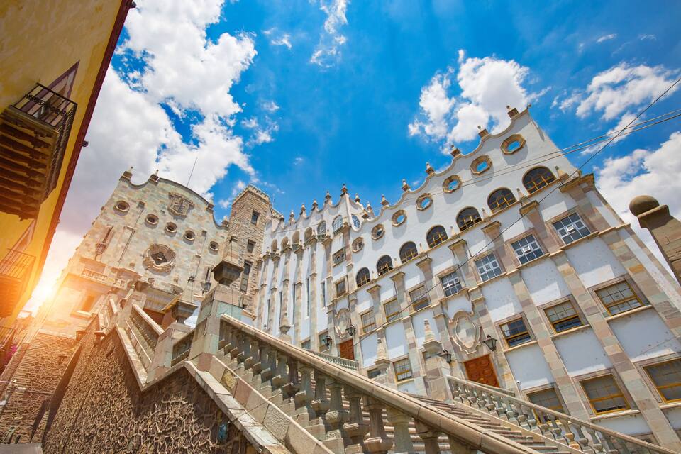 Um edifício histórico com arquitetura elaborada, uma escadaria imponente e um céu azul claro com nuvens.