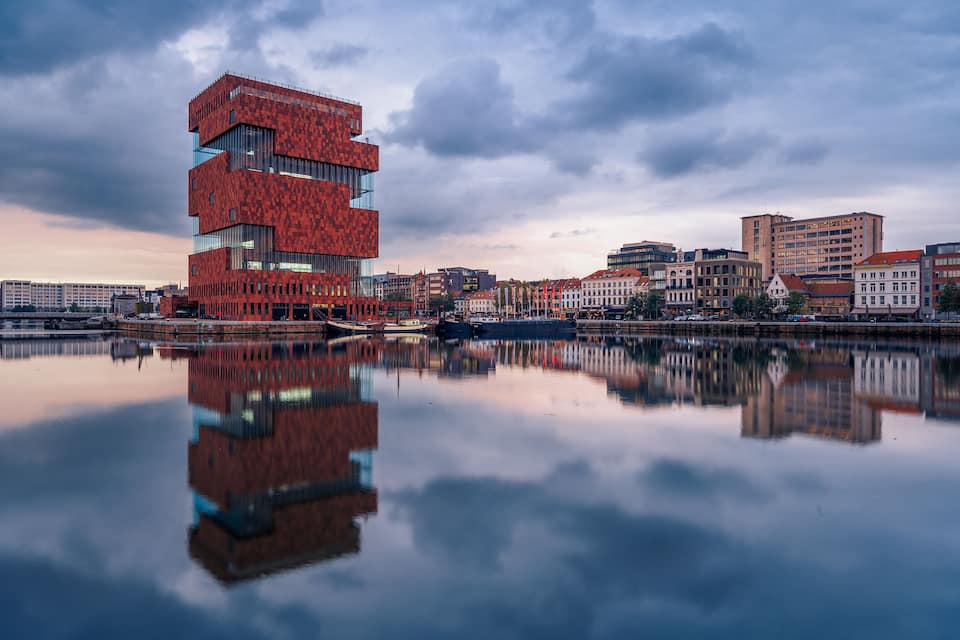 Um edifício vermelho moderno com design exclusivo, refletindo em águas calmas, rodeado por outros edifícios.