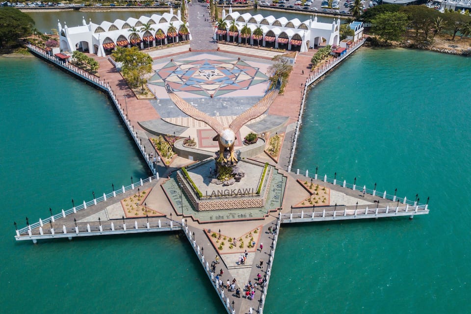 Uma grande ponte com múltiplos arcos e uma ilha central com uma imponente estátua de águia e a palavra 'LANGKAWI' escrita na ilha.