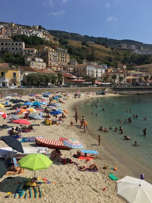 Praia cheia, com pessoas nadando e tomando sol, cercada por guarda-sóis e cadeiras de praia coloridas.