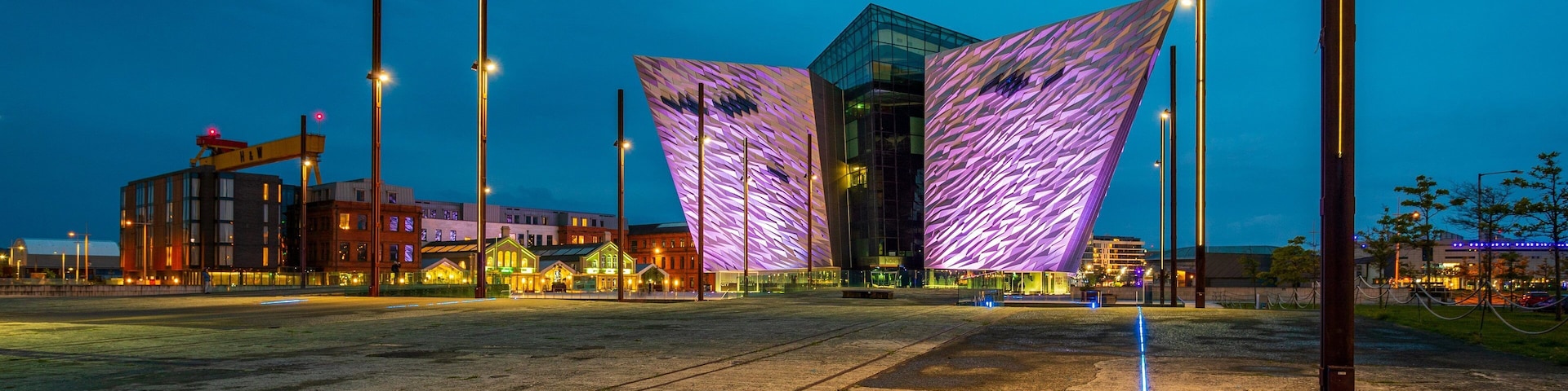 Um edifício moderno com fachada iluminada em roxo e uma ampla praça com linha férrea.