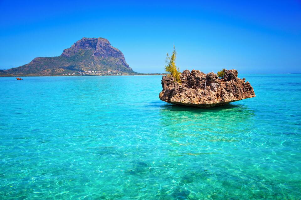 Mar azul e límpido com um afloramento rochoso em primeiro plano e uma montanha ao fundo.