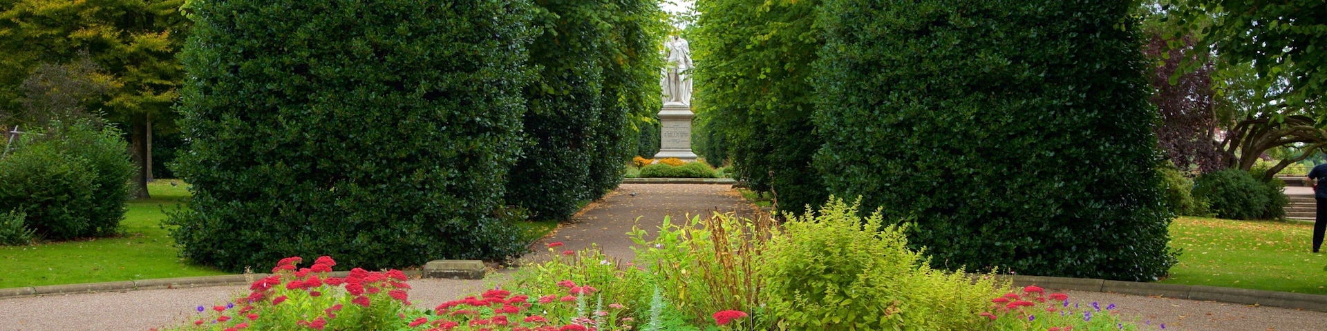 Um jardim formal com uma estátua central, cercado por sebes bem aparadas e canteiros floridos vibrantes.