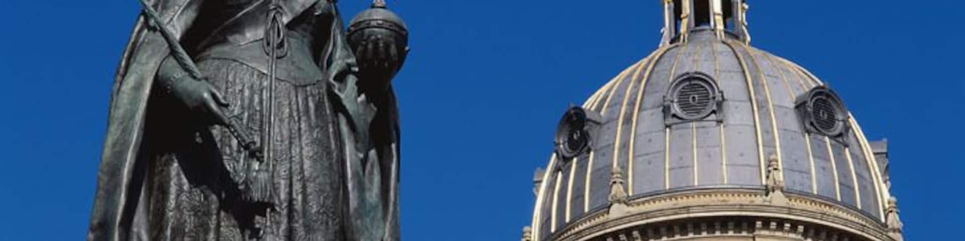 Estátua de mulher segurando uma cruz e um livro, em frente a uma grande cúpula com cruz no topo.