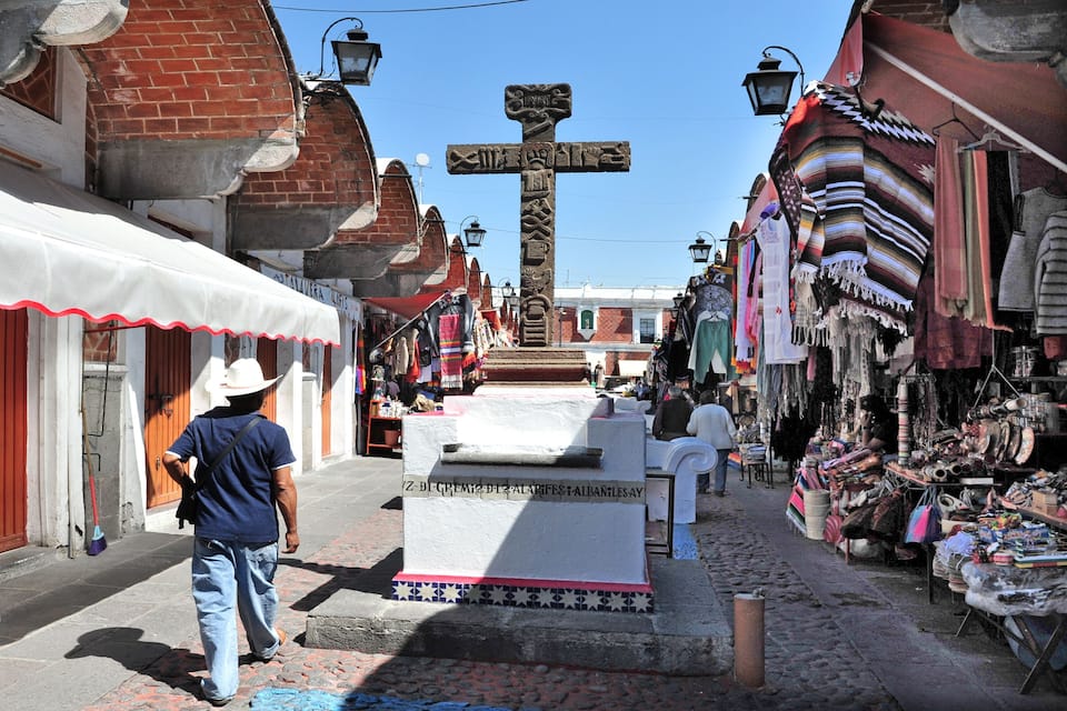 Uma rua de calçada com um grande monumento em forma de cruz, lojas e tecidos tradicionais.
