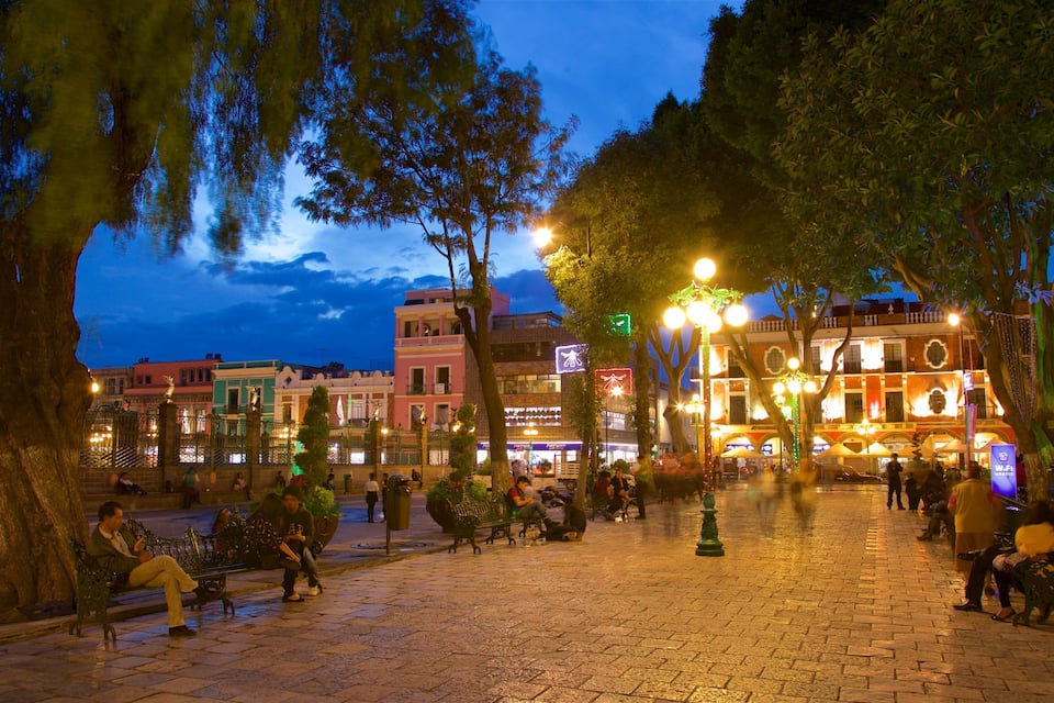 Uma praça à noite com pessoas sentadas em bancos, árvores e edifícios iluminados.