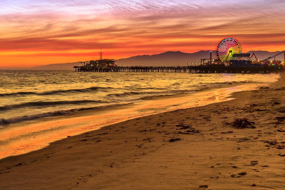 Uma praia ao pôr do sol, com um píer e parque de diversões ao fundo.