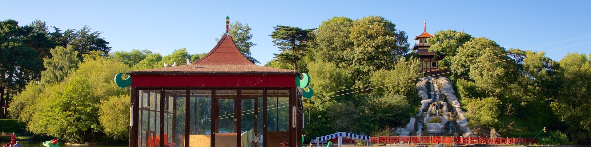 Um pavilhão com cobertura de vidro à beira de um lago, um barco verde e uma estrutura semelhante a um pagode ao fundo.