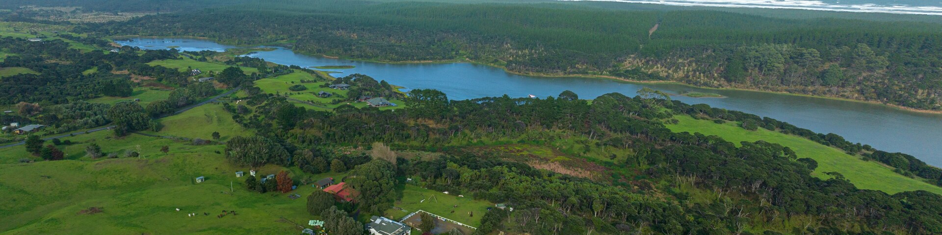 Paisagem costeira com uma grande extensão de água, campos verdes e um pequeno povoado com casas e uma estrada.