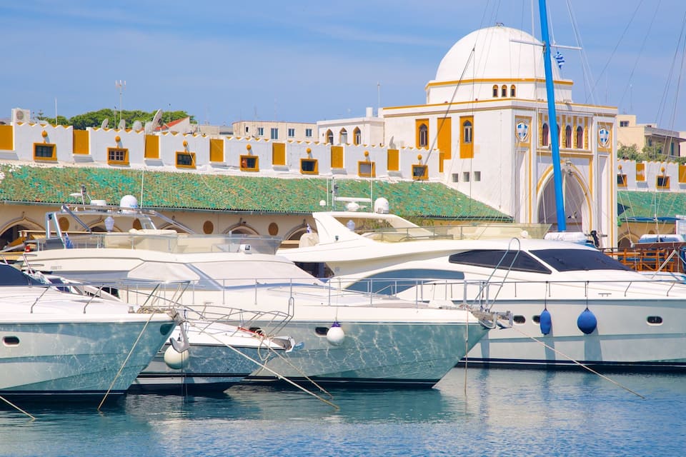 Uma marina com vários iates atracados, um edifício branco com cúpula e uma estrutura com telhado verde ao fundo.