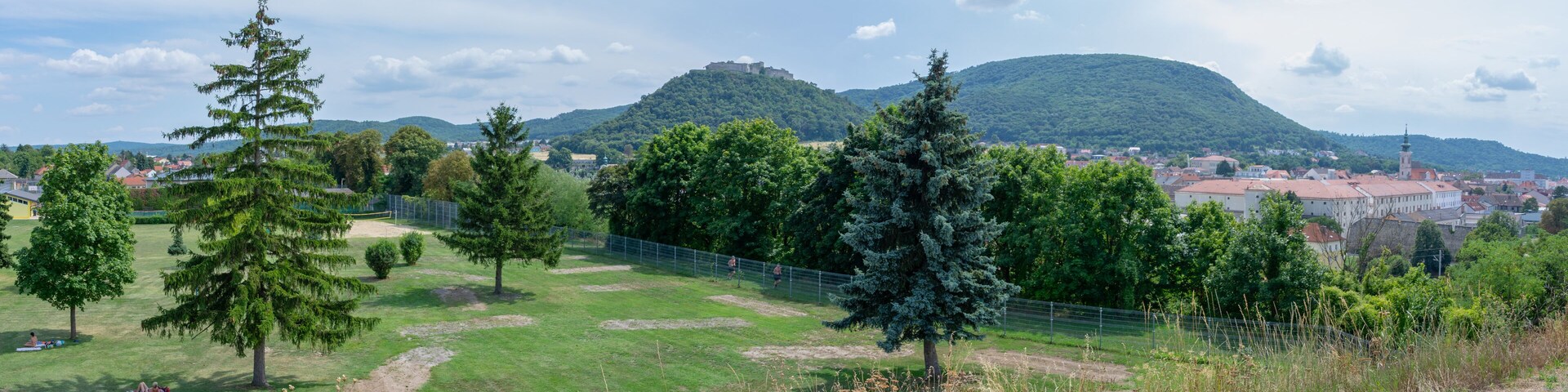 Um parque com árvores, uma área cercada e uma colina com um castelo ao fundo.