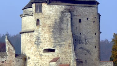 Uma torre de pedra medieval com telhado cônico, rodeada por uma muralha fortificada e um edifício menor com telhado vermelho.