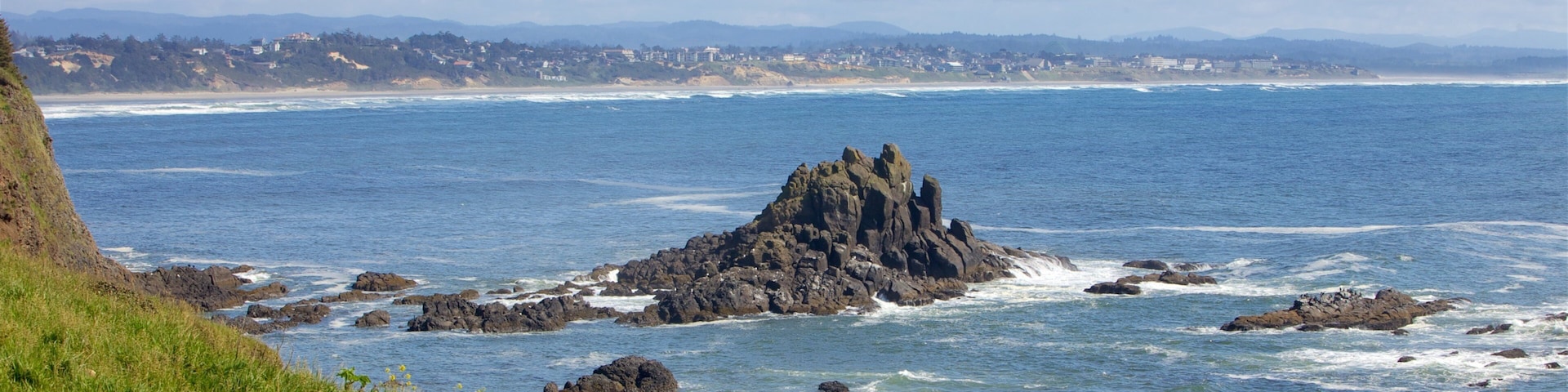 Litoral rochoso com uma formação rochosa imponente ao centro, cercada por ondas do oceano e um horizonte distante com edifícios.