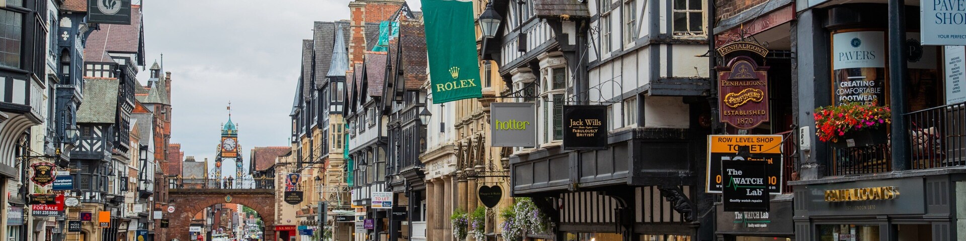 Uma rua movimentada em uma cidade histórica, com edifícios em estilo Tudor, lojas e uma imponente torre do relógio.