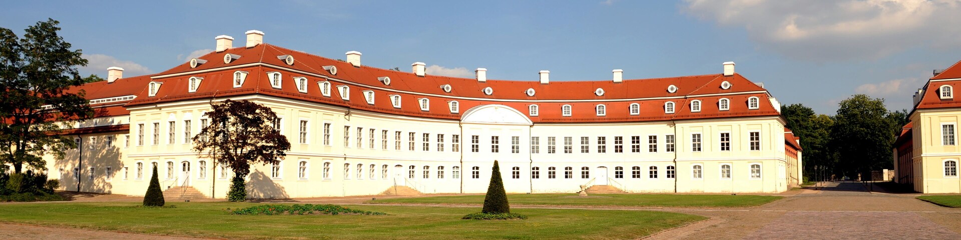 Um edifício grande e histórico com telhado vermelho e paredes brancas, rodeado por um pátio pavimentado e algumas árvores.