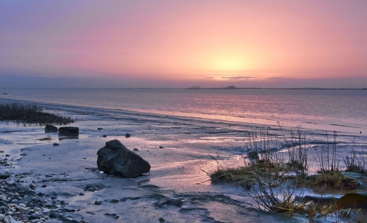 Uma praia ao pôr do sol com rochas e vegetação rasteira.