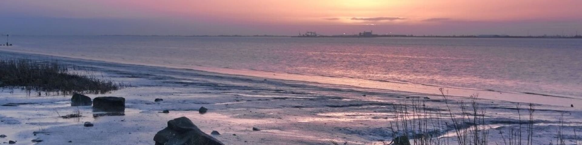 Uma praia ao pôr do sol com rochas e vegetação rasteira.