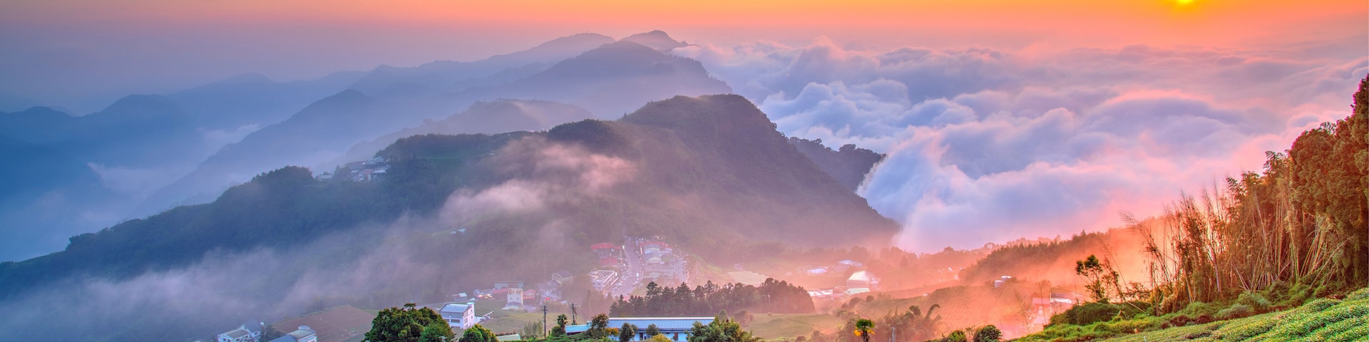 Nascer do sol sobre uma paisagem montanhosa com plantações de chá em terraços e uma aldeia aninhada no vale.