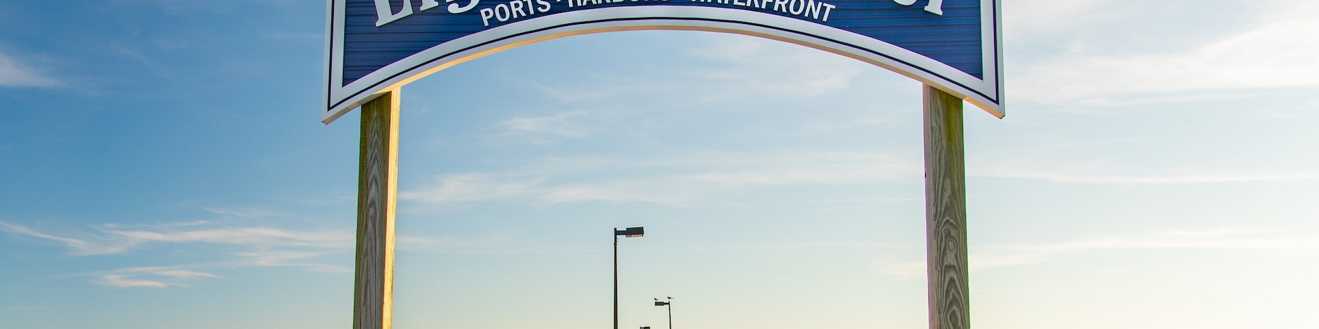 Um cais de madeira leva a uma placa com a inscrição 'Lighthouse Pier', com um céu claro e águas calmas ao fundo.