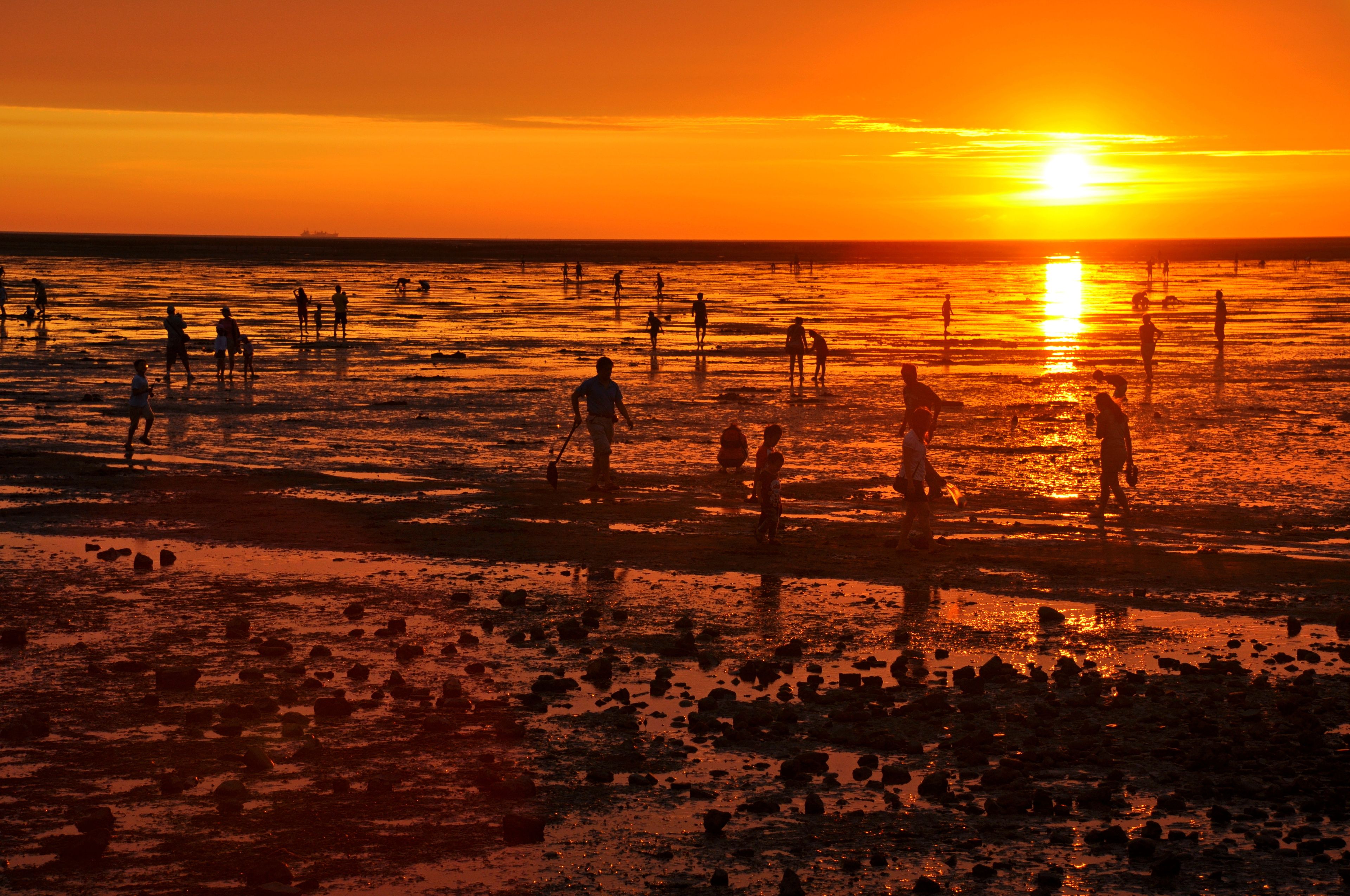 Um pôr do sol sobre uma praia, com pessoas caminhando e brincando na água.
