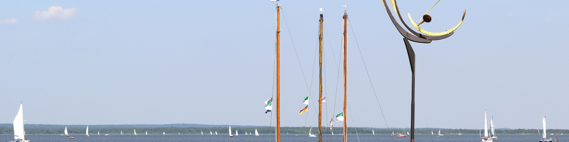Um lago calmo com vários barcos atracados no cais, cercado por vegetação e veleiros ao longe.