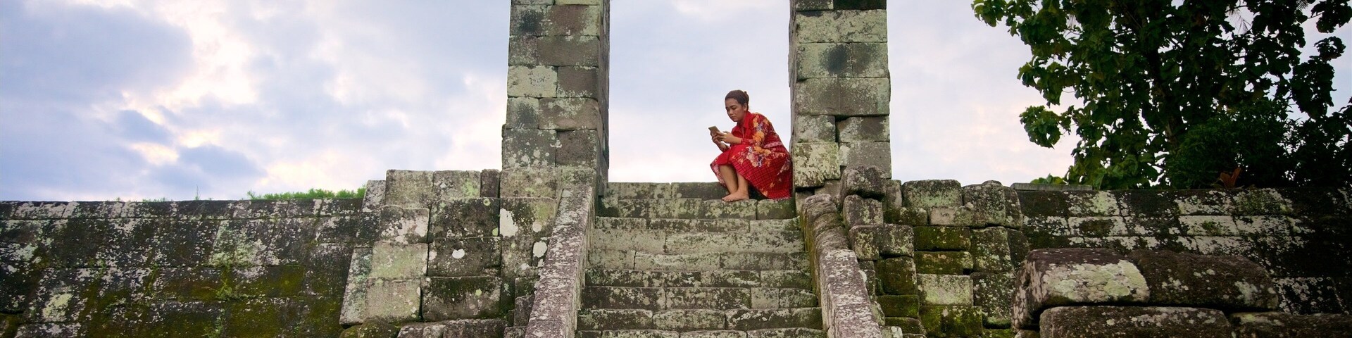 Mulher de vestido vermelho sentada em degraus de pedra antigos, com paredes cobertas de musgo e uma palmeira ao fundo.