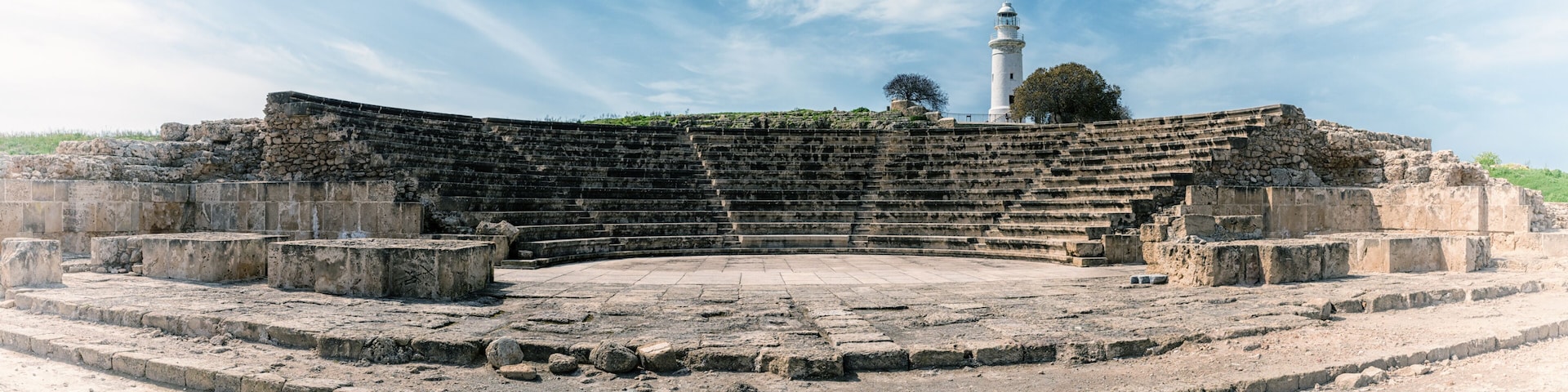 Um antigo anfiteatro com degraus de pedra e palco central, rodeado por ruínas e um farol ao fundo.
