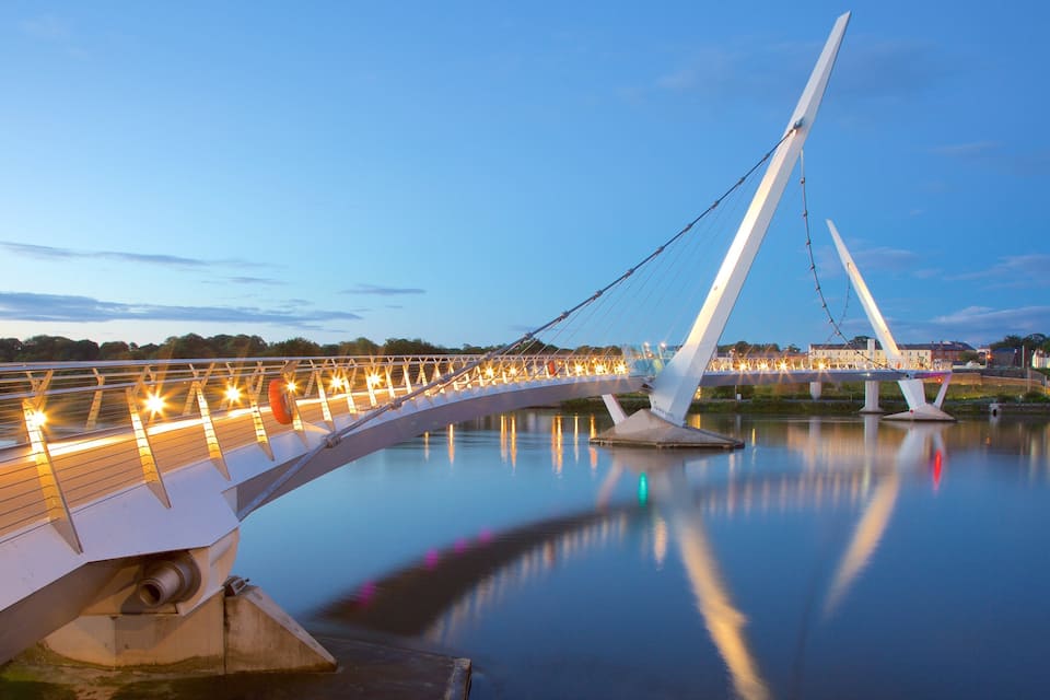 Uma ponte moderna com luzes iluminadas ao entardecer sobre um rio calmo, refletindo a estrutura.