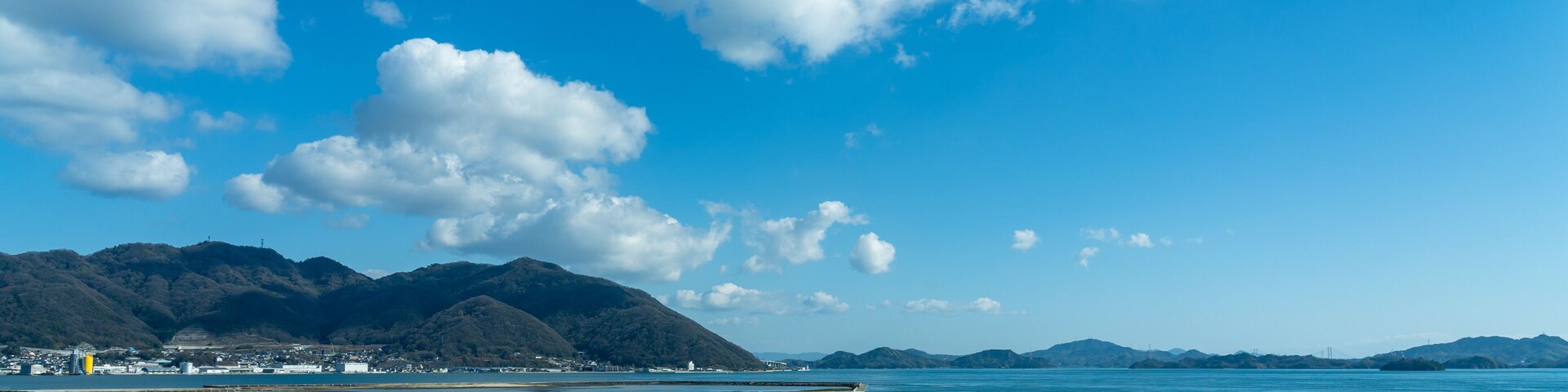 Paisagem costeira com céu azul límpido, mar calmo e uma cordilheira ao fundo.