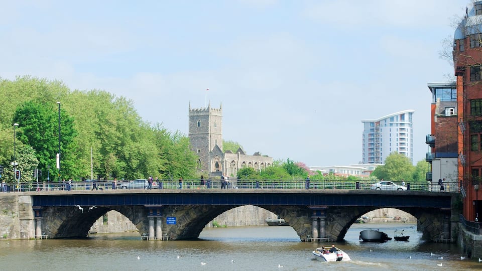 Uma ponte de pedra sobre um rio, com barcos e cisnes, uma torre de igreja e edifícios modernos ao fundo.
