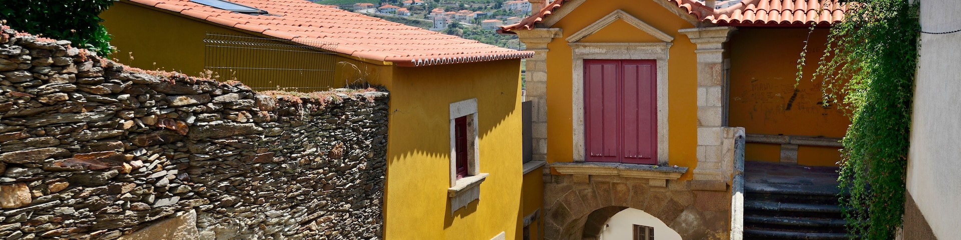 Uma viela estreita com uma parede de pedra, um edifício amarelo com porta vermelha e uma escadaria que leva a uma igreja com uma cruz no topo.