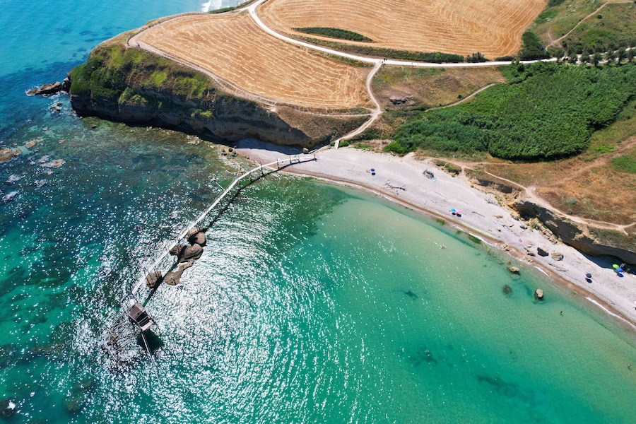 Vista aérea de uma área costeira com uma praia clara, falésias rochosas e uma ponte ligando a terra ao mar.