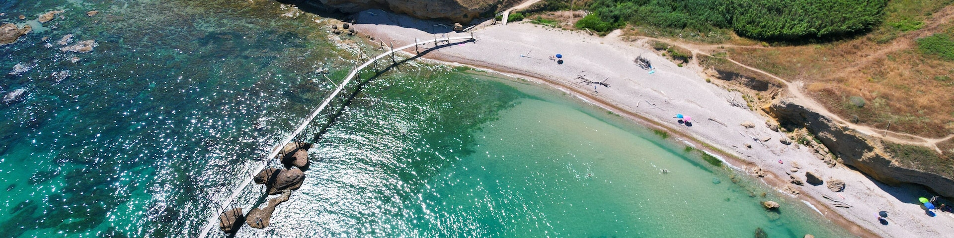 Vista aérea de uma área costeira com uma praia clara, falésias rochosas e uma ponte ligando a terra ao mar.