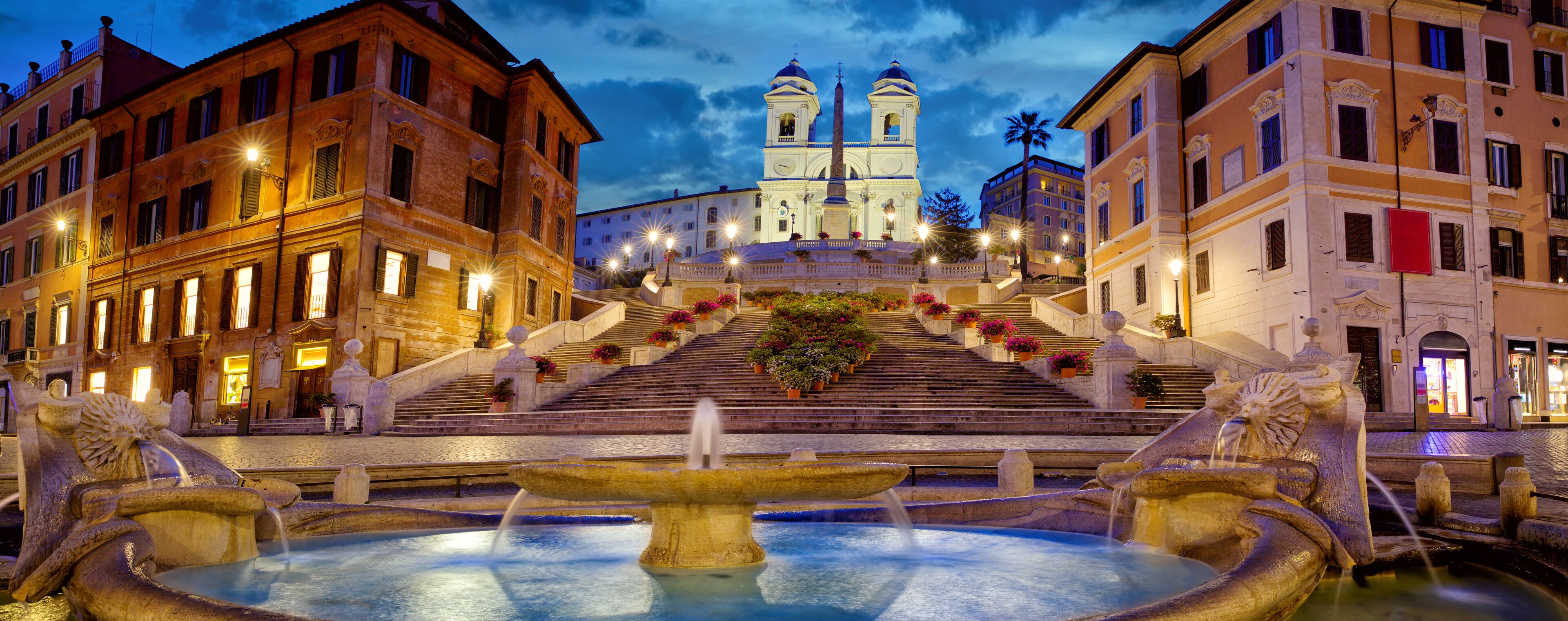 Uma escadaria imponente leva a uma catedral com duas torres, rodeada por edifícios bem iluminados e uma fonte em primeiro plano.