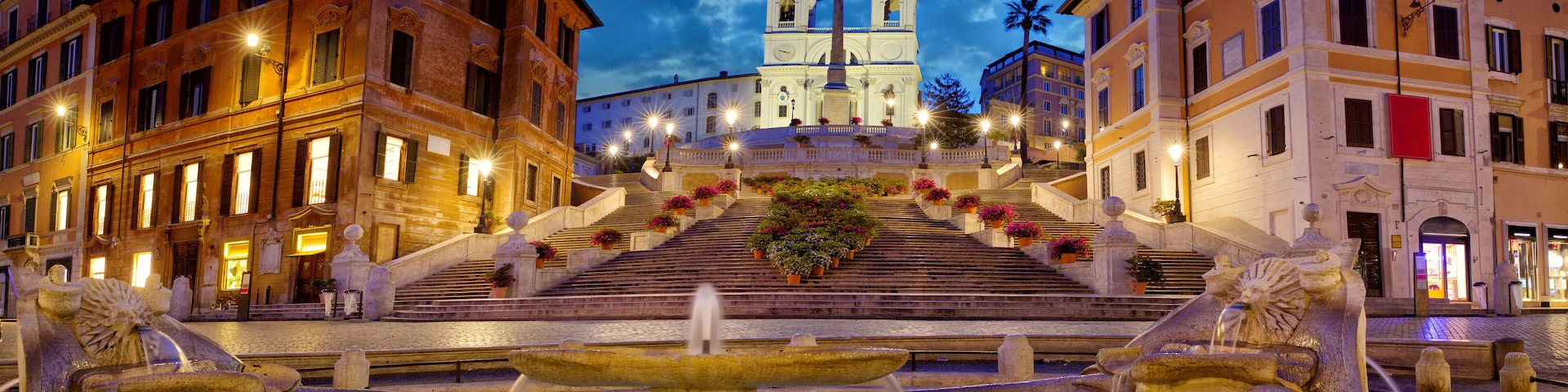 Uma escadaria imponente leva a uma catedral com duas torres, rodeada por edifícios bem iluminados e uma fonte em primeiro plano.