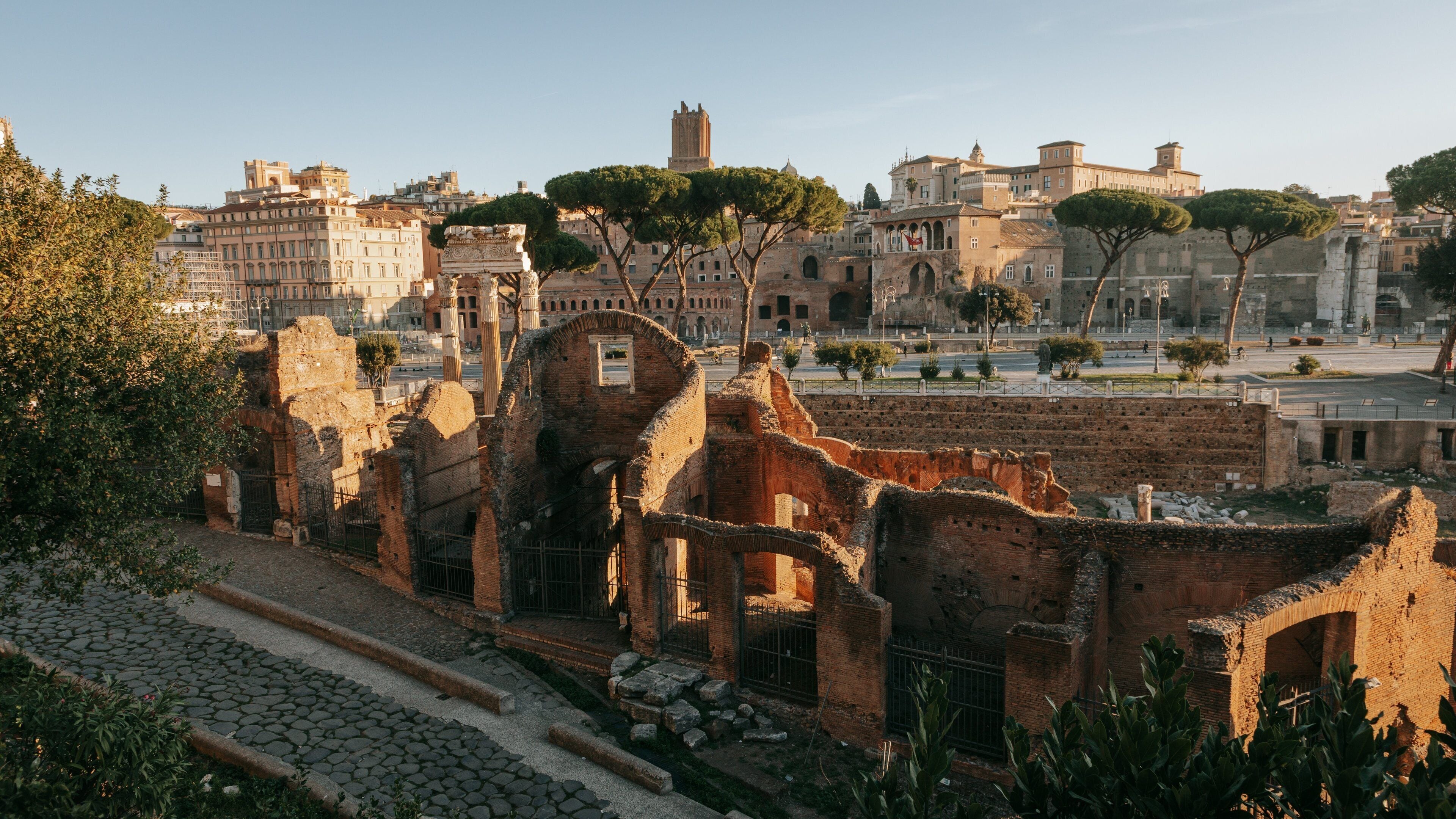 Ruínas antigas com arcos e colunas, rodeadas por árvores e edifícios históricos.