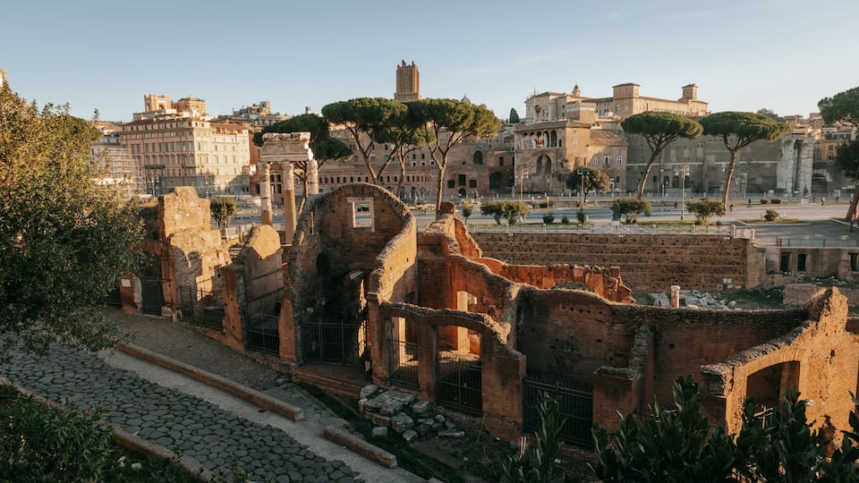 Ruínas antigas com arcos e colunas, rodeadas por árvores e edifícios históricos.