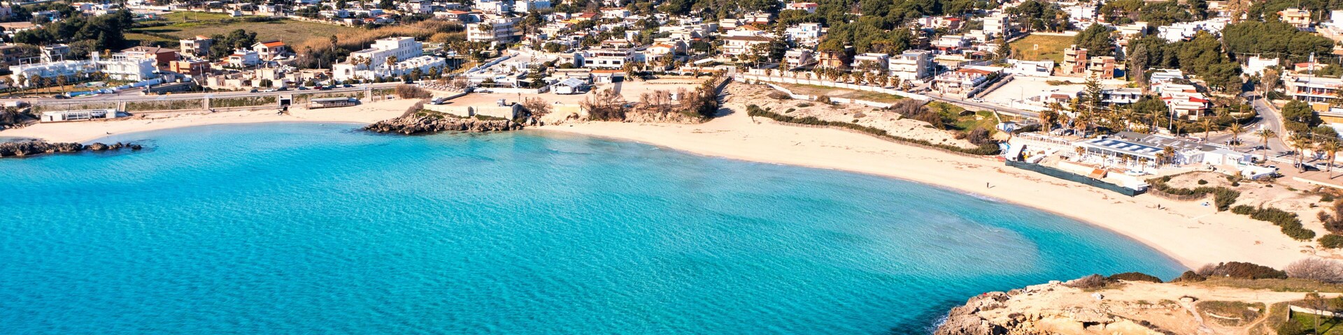 Uma vila costeira com praia de areia, águas azuis cristalinas e uma mistura de edifícios residenciais e comerciais.