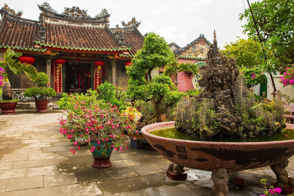 Um templo tradicional com arquitetura ornamentada, uma grande fonte de pedra e plantas floridas vibrantes.