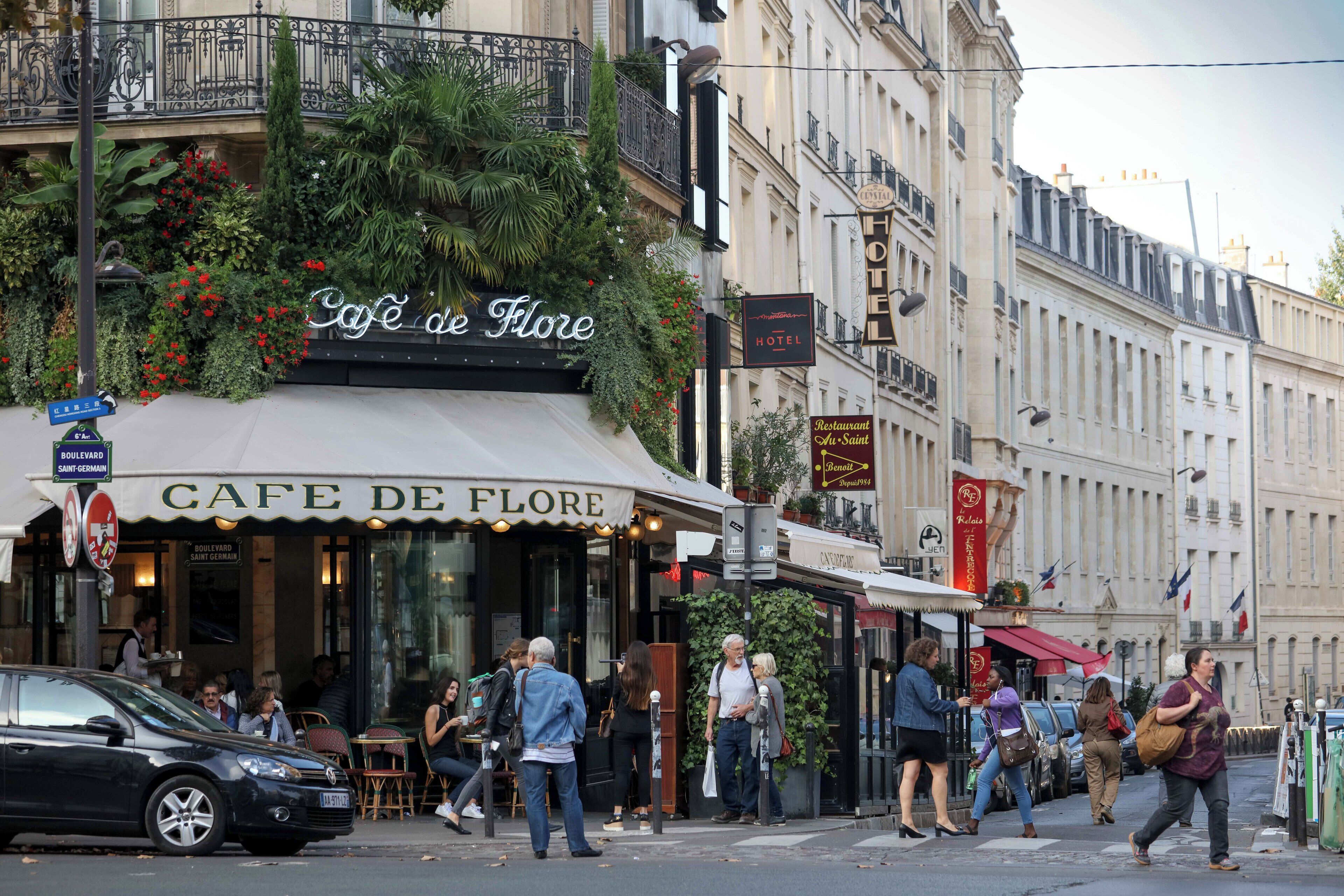 Um café chamado Café de Flore com mesas ao ar livre, rodeado por edifícios e pedestres.