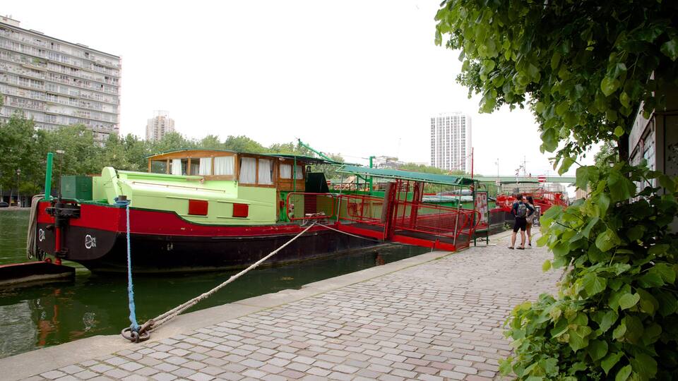 Um barco verde e vermelho atracado em um canal, com um passeio de tijolos e edifícios ao fundo.