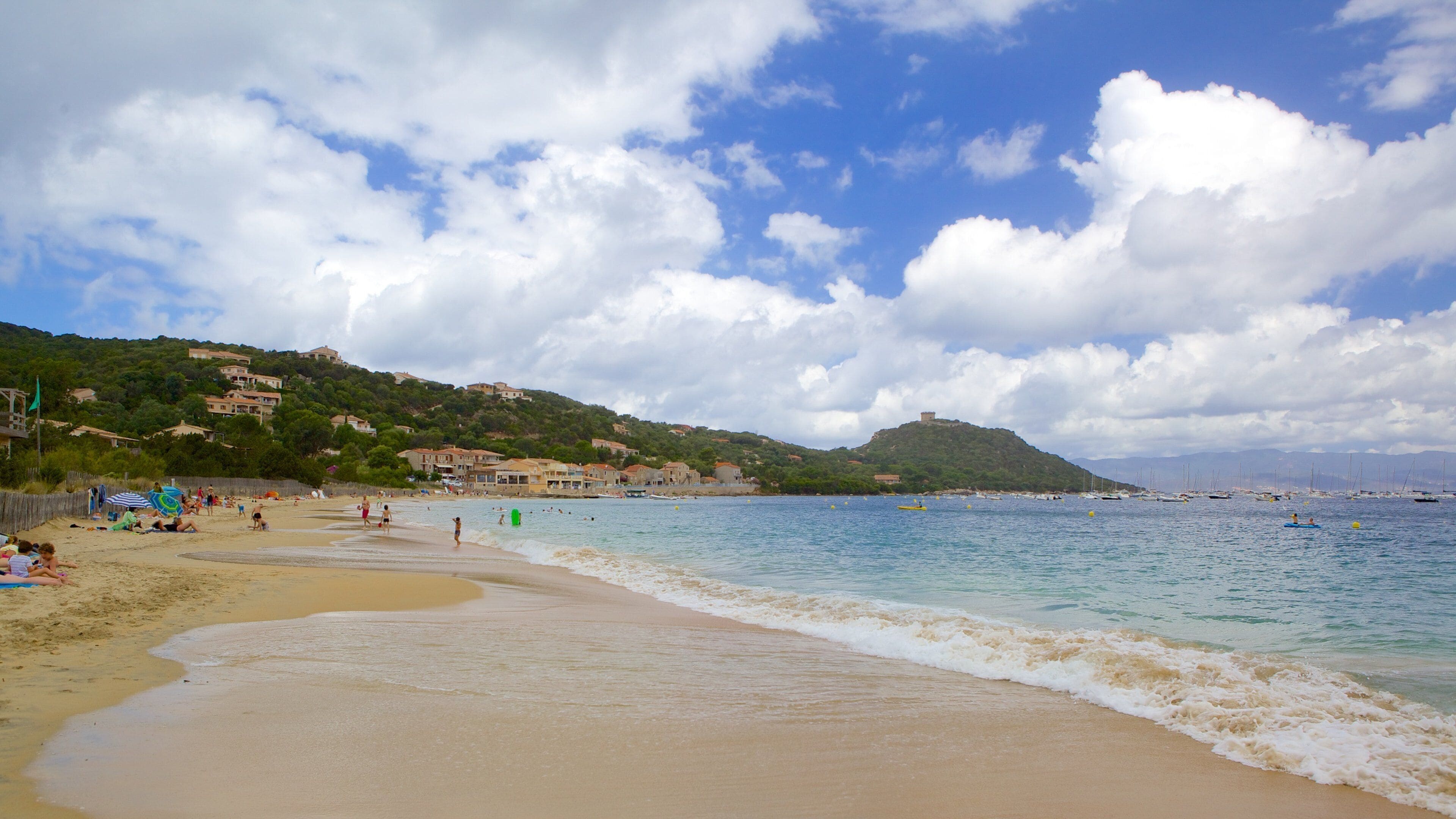 Uma praia com águas cristalinas, areal dourado e edifícios numa encosta ao fundo.