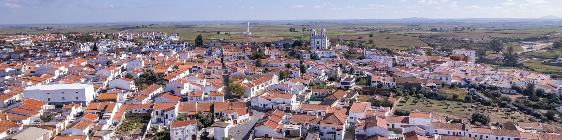 Uma cidade com telhados vermelhos e edifícios brancos, uma igreja com campanário e um farol ao longe.