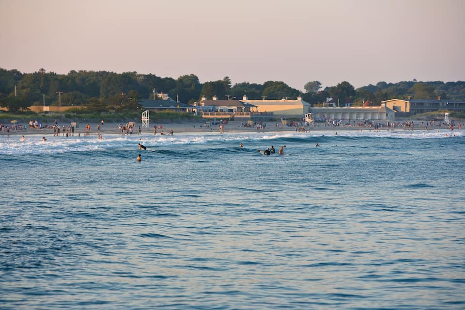 Uma praia com pessoas nadando e praticando surf, edifícios ao fundo e uma colina coberta de árvores.