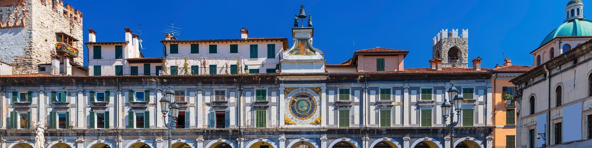 Uma praça histórica com uma torre do relógio imponente, arcos e uma estátua.