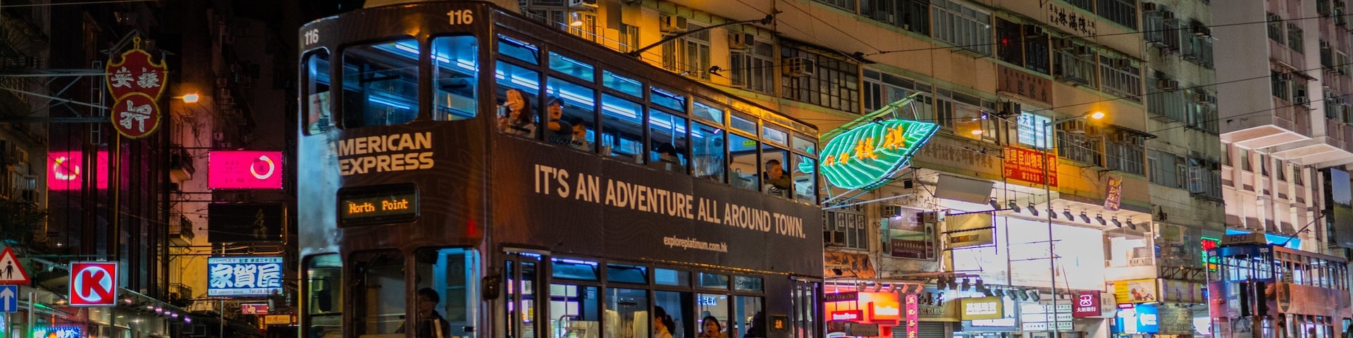 Um ônibus de dois andares com 'American Express' e a inscrição 'É uma aventura por toda a cidade' nas laterais, dirigindo em uma rua da cidade à noite.