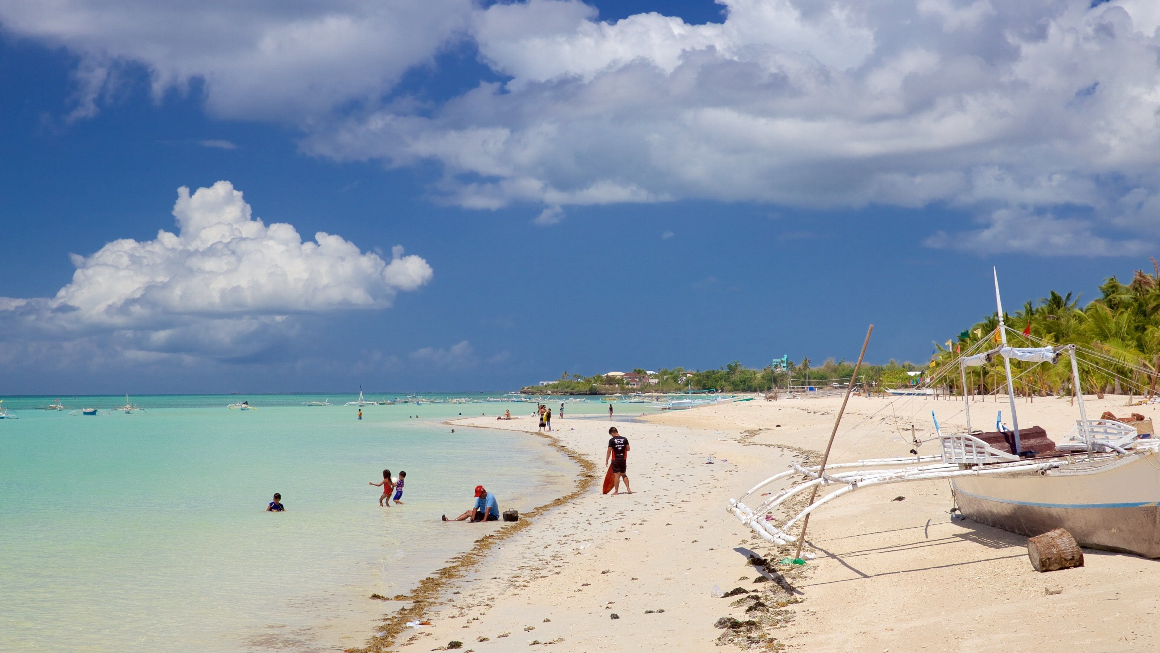 Uma praia com águas turquesas cristalinas, pessoas a nadar e passear, e um barco na areia.