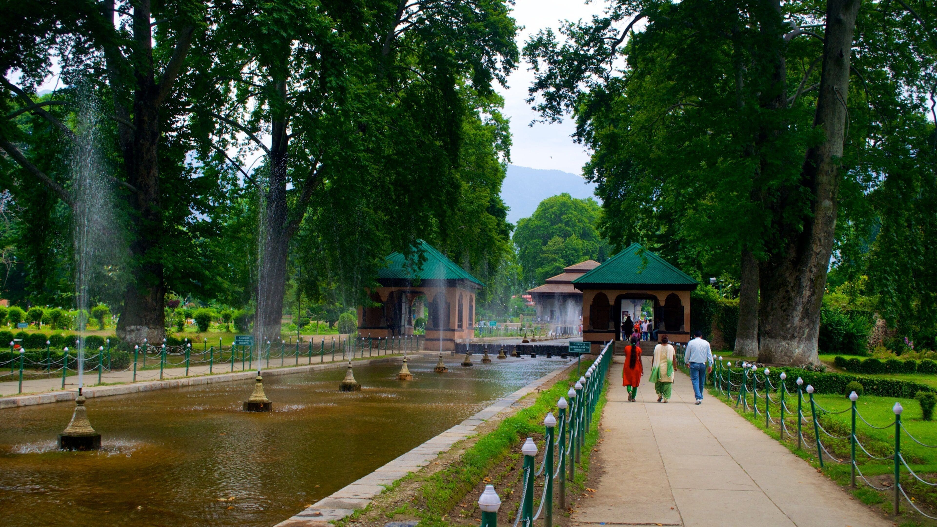 Um parque com um espelho d'água, dois pavilhões com telhado verde e um caminho ladeado por uma cerca.